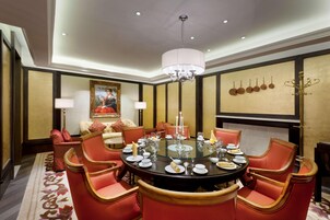 Uma sala de jantar formal com uma mesa redonda, cadeiras estofadas em vermelho, um candelabro e um quadro grande na parede.