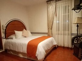 Quarto de hotel com cama de cabeceira em madeira, roupa de cama branca, manta laranja, uma cadeira e um abajur.