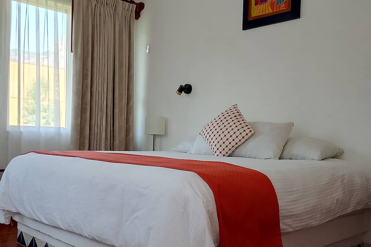 Cama bem arrumada com manta vermelha, almofada estampada e luminária fixada na parede.