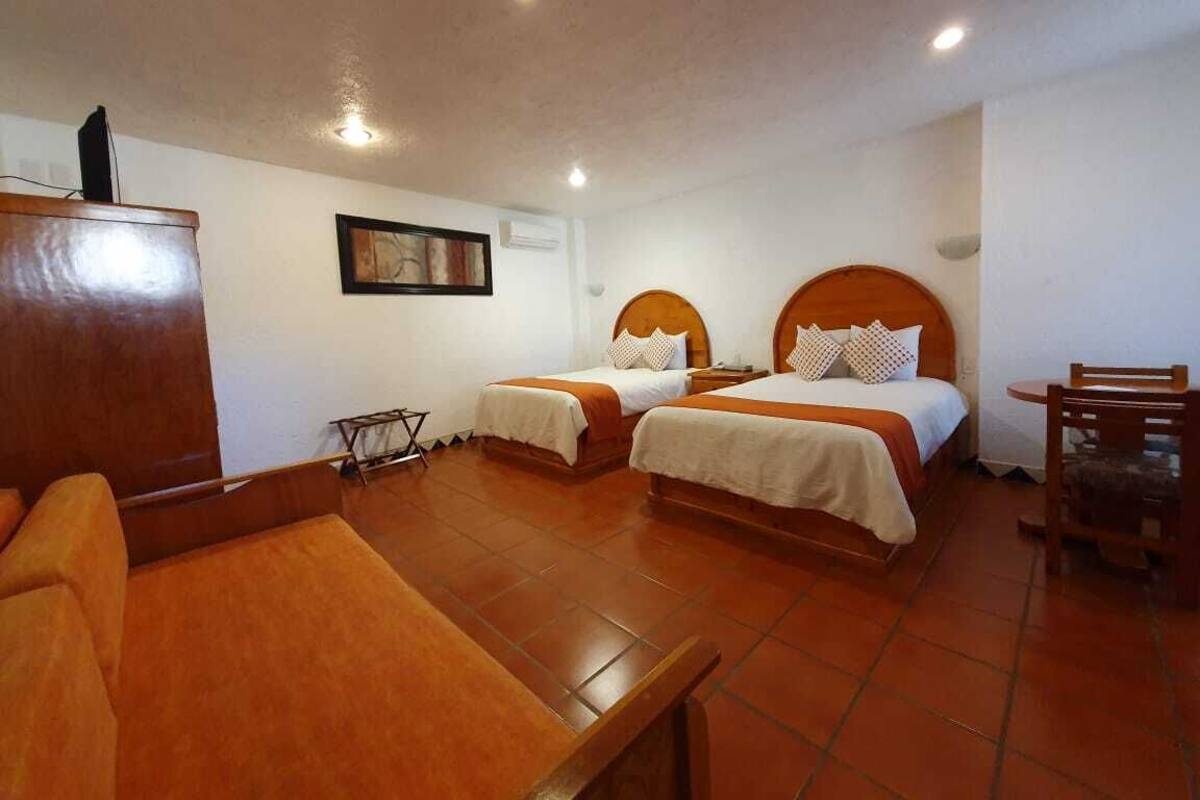 Quarto de hotel com duas camas, uma escrivaninha de madeira, uma cadeira e uma mesinha.