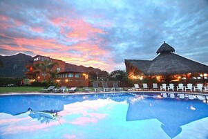 Piscina de resort com espreguiçadeiras, uma estrutura de palha e montanhas ao fundo durante o pôr do sol.