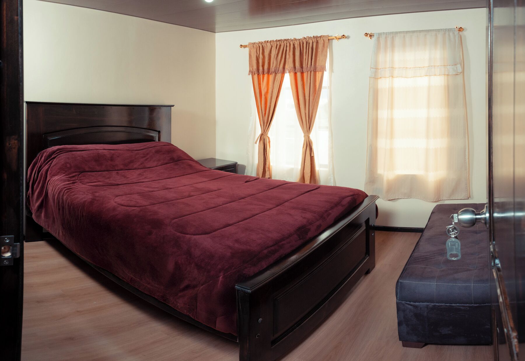 Quarto com cama, mesa de cabeceira, um banco e duas janelas com cortinas.