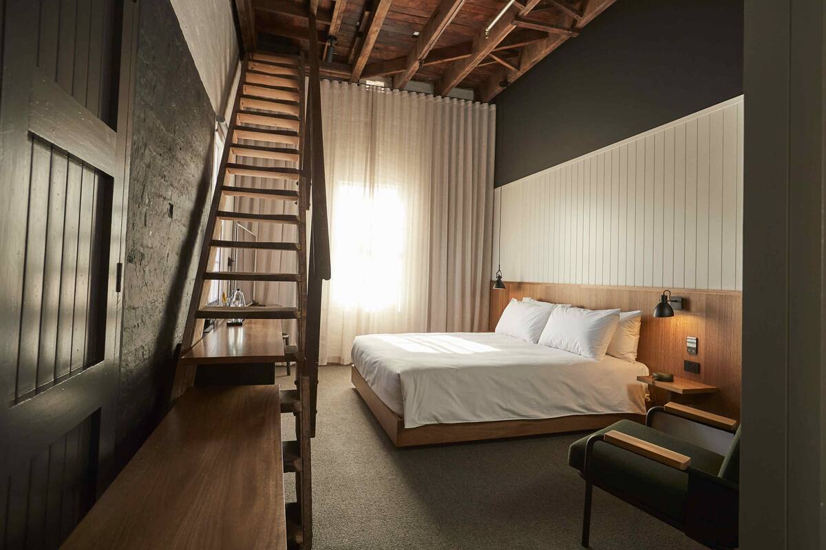 Quarto com cama de madeira, mesa de cabeceira, abajur e escada.