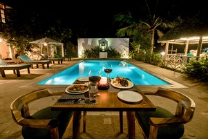 Uma área de refeições ao ar livre com piscina, cadeiras de madeira, uma mesa posta para dois e uma estátua de Buda ao fundo.