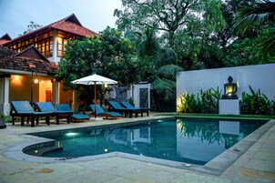 Piscina com espreguiçadeiras e uma estátua, rodeada por vegetação exuberante e um edifício com telhado vermelho.