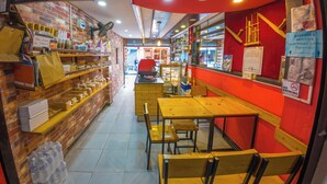Um café estreito com paredes de tijolo, mesas e cadeiras de madeira, um balcão com diversos itens e uma parede vermelha com placas.