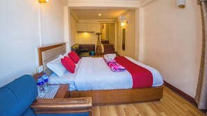 Quarto de hotel com cama, um banquinho de madeira, mesa com uma garrafa de água e uma cadeira.