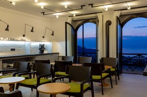 Uma área de refeições moderna com mesas e cadeiras redondas, um bar com bancada branca e grandes janelas em arco que oferecem vista para o mar.