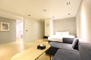 Quarto de hotel moderno com uma cama, mesa de cabeceira com controle remoto, criado-mudo pequeno e área de estar com um banco.