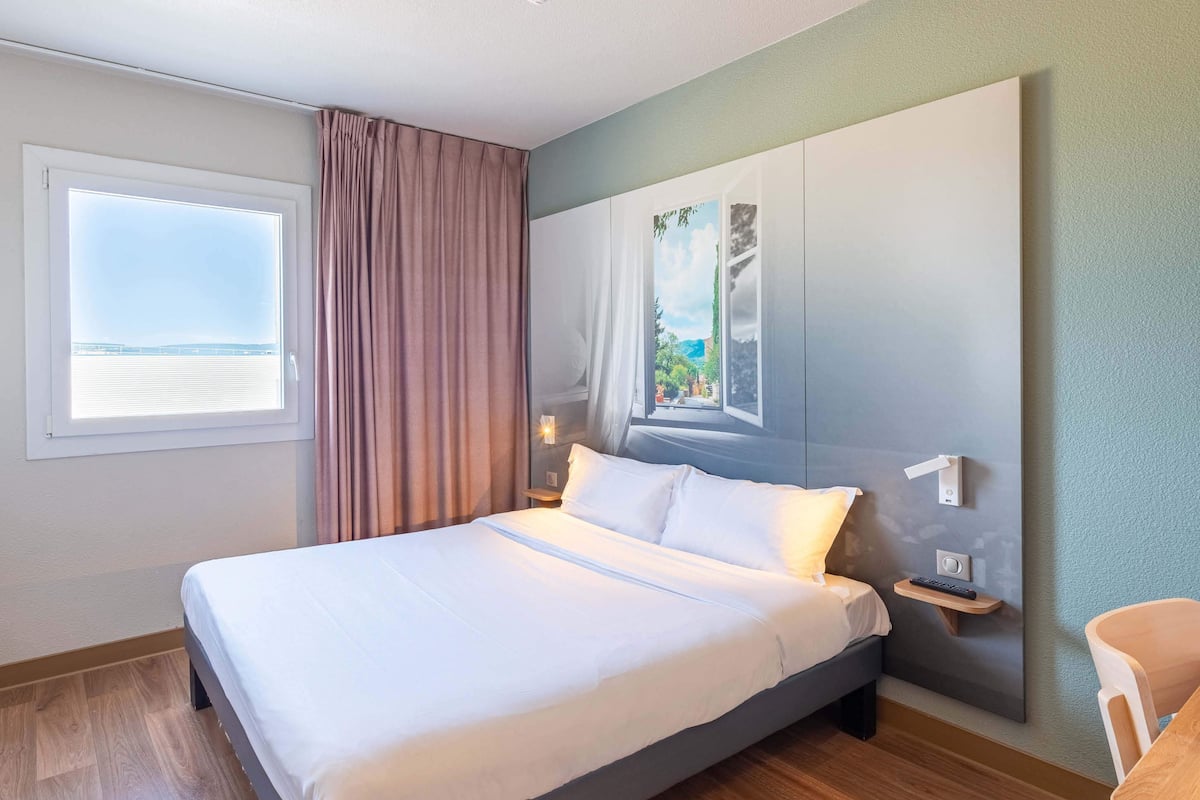 Quarto de hotel com uma cama, janela com cortinas, mesa de cabeceira e prateleira fixa na parede com controle remoto.