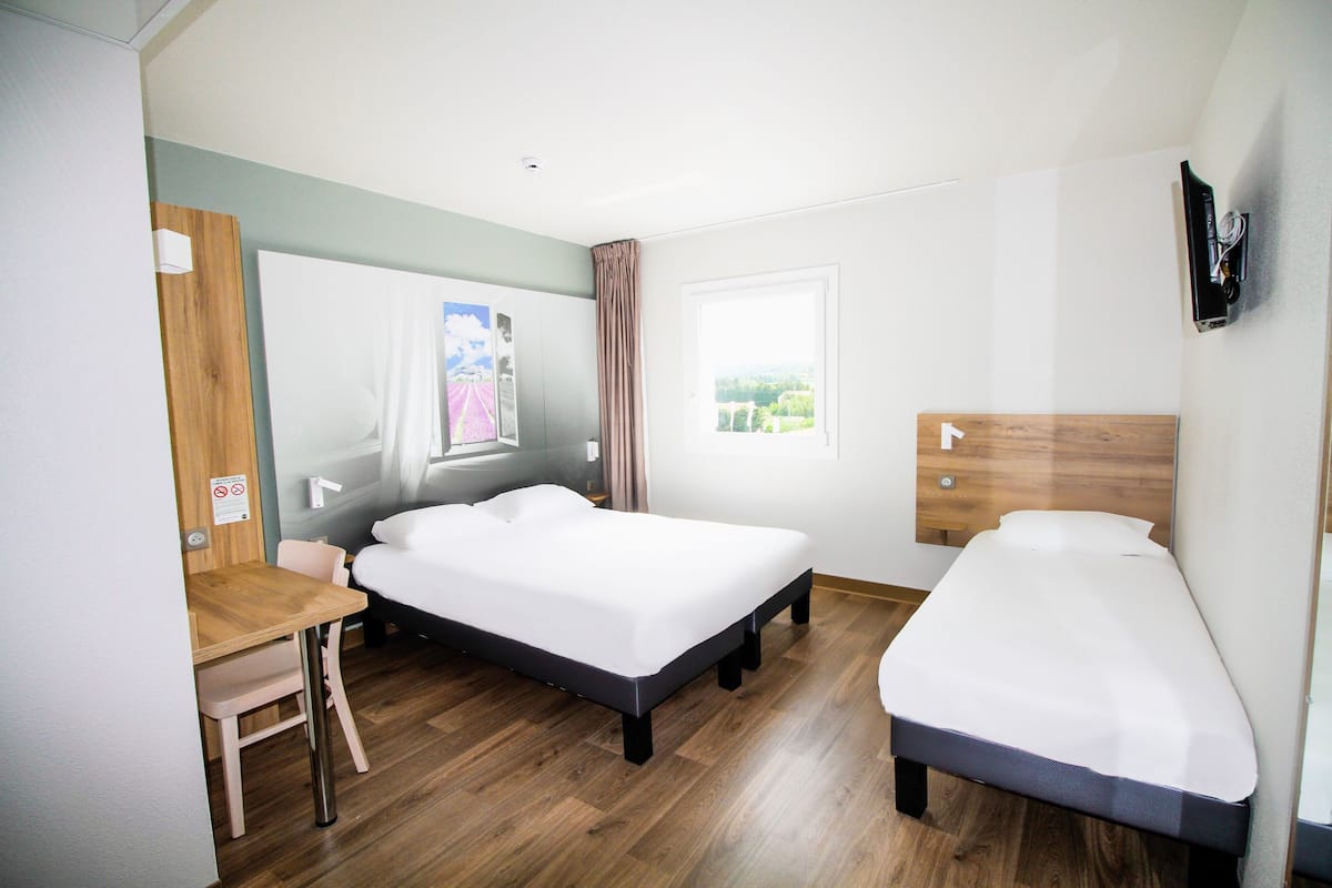 Quarto de hotel com duas camas de solteiro, uma escrivaninha de madeira, uma cadeira, janela com cortinas e televisão na parede.