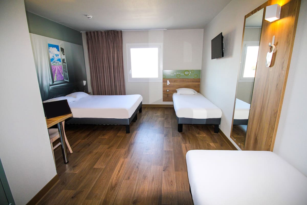 Quarto de hotel com duas camas de solteiro, uma escrivaninha, uma cadeira, um espelho e uma janela.