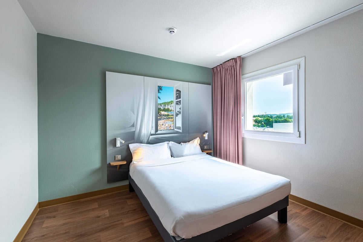 Quarto com cama, mesa de cabeceira, janela com cortinas e um quadro na parede.