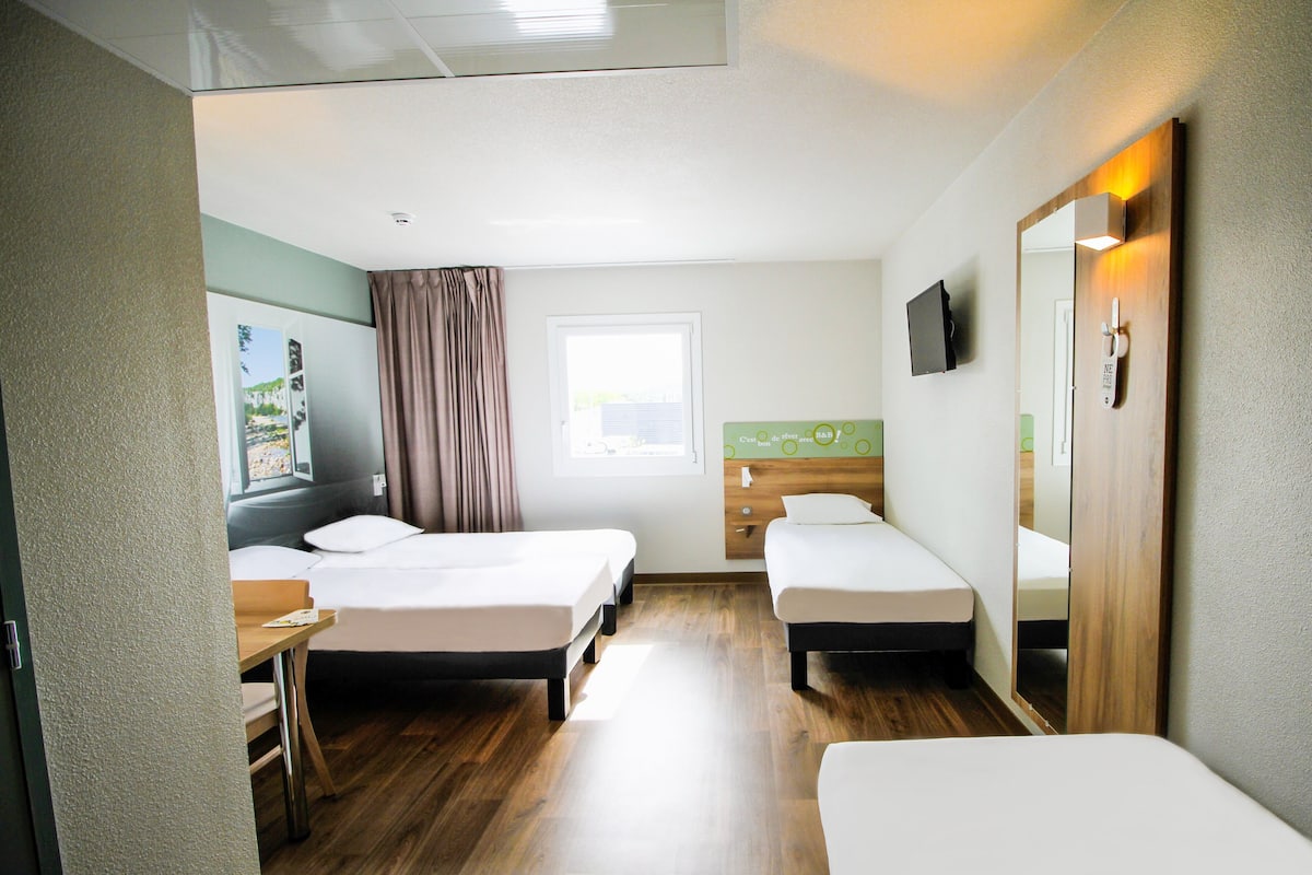 Quarto de hotel com duas camas de solteiro, uma escrivaninha, uma cadeira, uma janela com cortinas, uma TV de tela plana na parede e uma porta de madeira com espelho.
