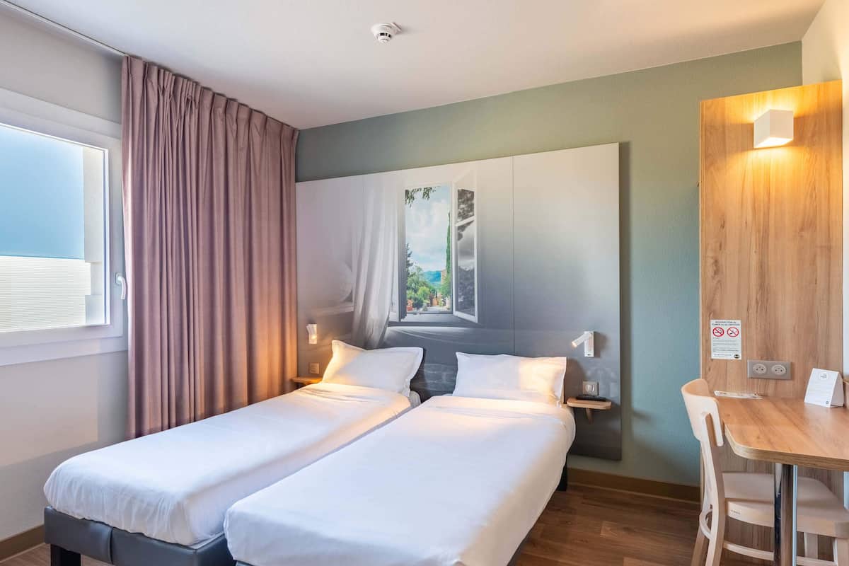 Quarto de hotel com duas camas de solteiro, uma secretária de madeira, uma cadeira, janela com cortinas e luminária fixada na parede.
