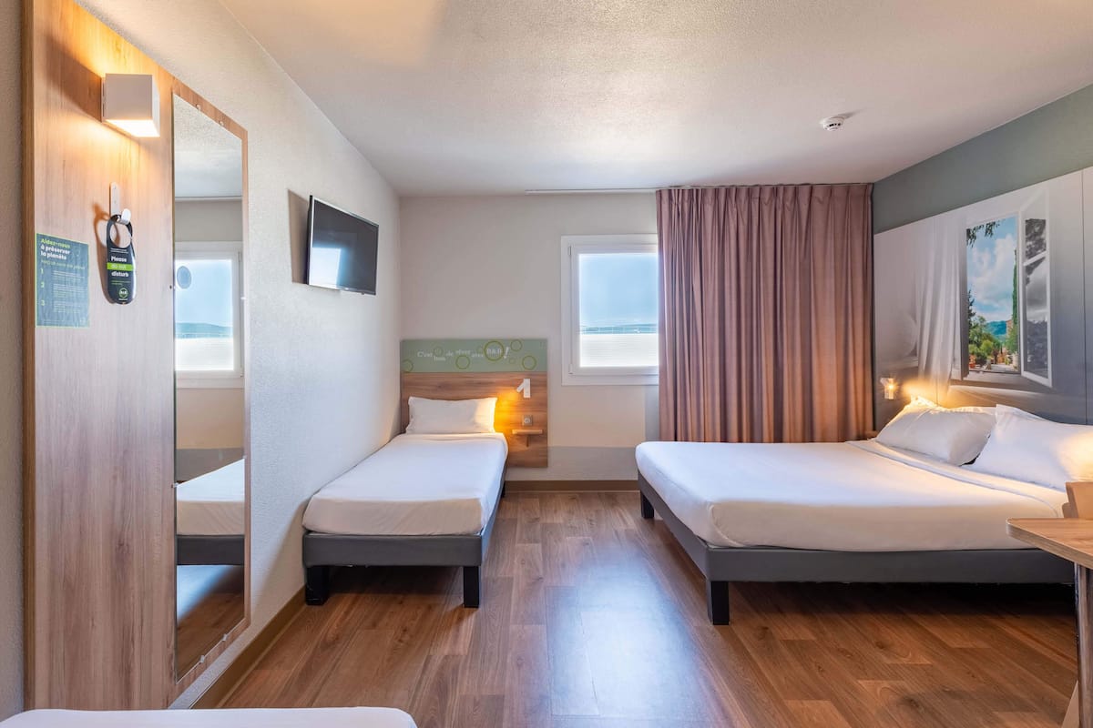 Quarto de hotel com duas camas de solteiro, piso de madeira, televisão, espelho e janela com cortinas.