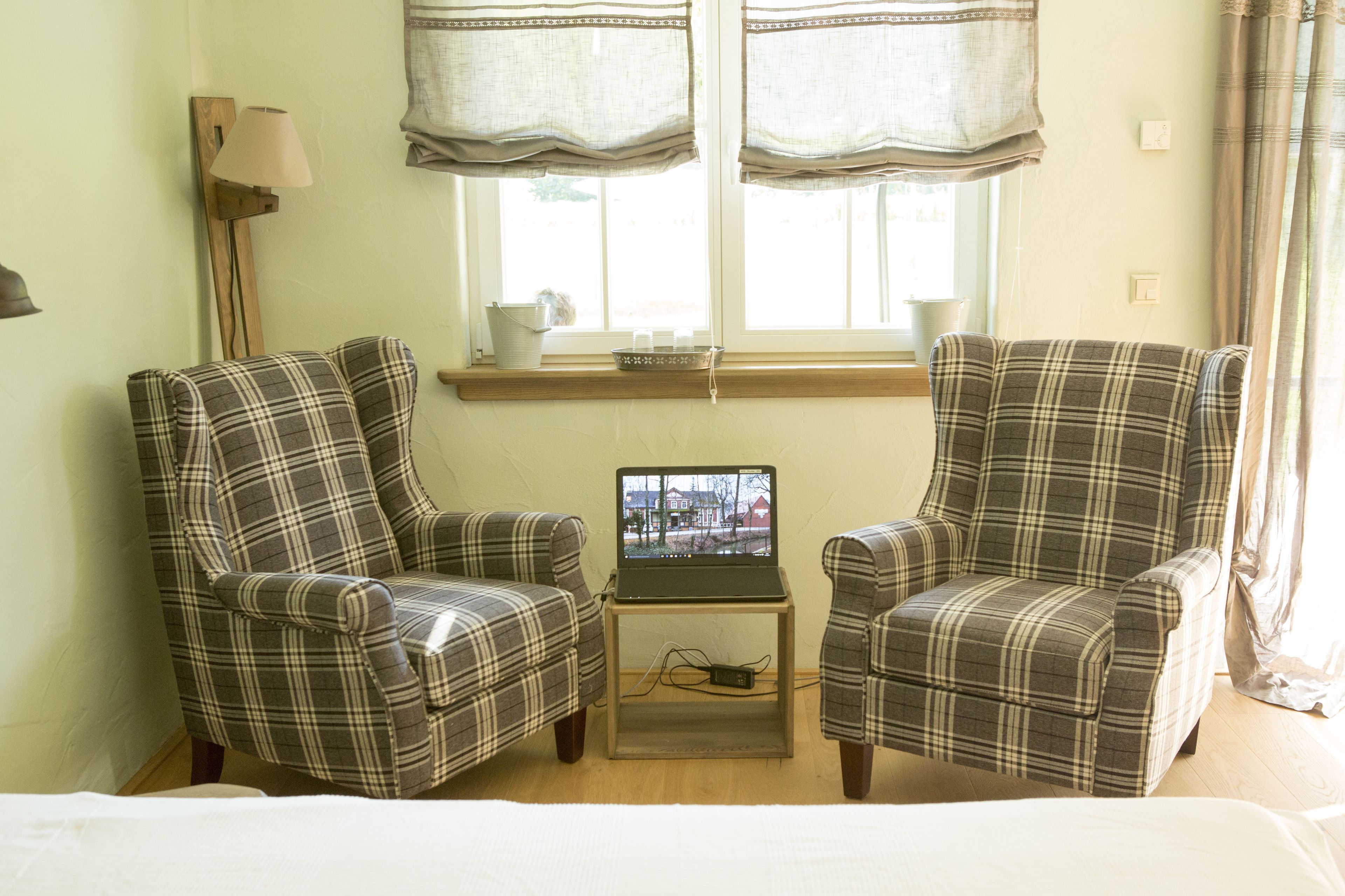 Quarto com duas poltronas xadrez, uma pequena mesa com um laptop, uma luminária e uma janela com cortinas.