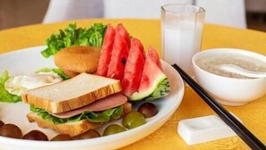Um prato com sanduíche, fatias de melancia, azeitonas e um copo de leite sobre uma toalha de mesa amarela.