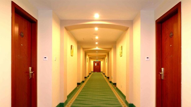 Corredor de hotel com carpete verde, portas de madeira e teto claro.