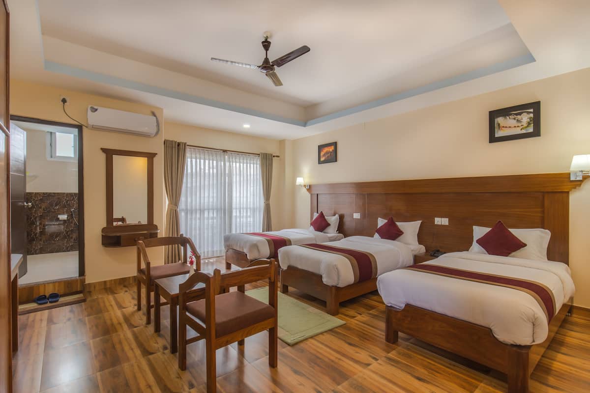 Quarto de hotel com duas camas, um espaço de estar com uma poltrona, piso de madeira, ventilador de teto e uma janela com cortinas.
