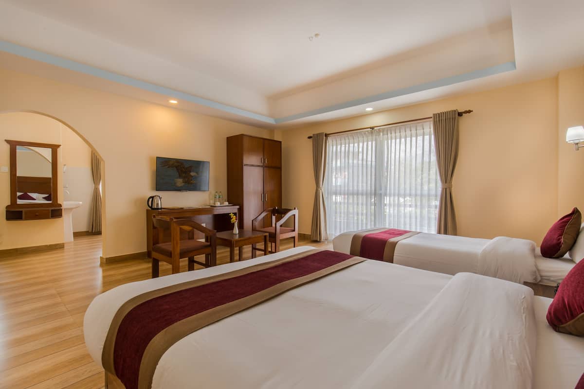 Quarto de hotel com duas camas, uma escrivaninha, uma cadeira e uma janela grande com cortinas.