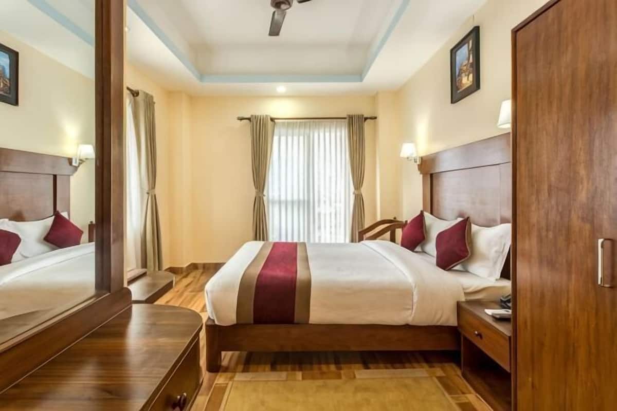 Quarto de hotel com duas camas, cabeceira de madeira, mesa de cabeceira, espelho, ventoinha no teto e janela com cortinas.