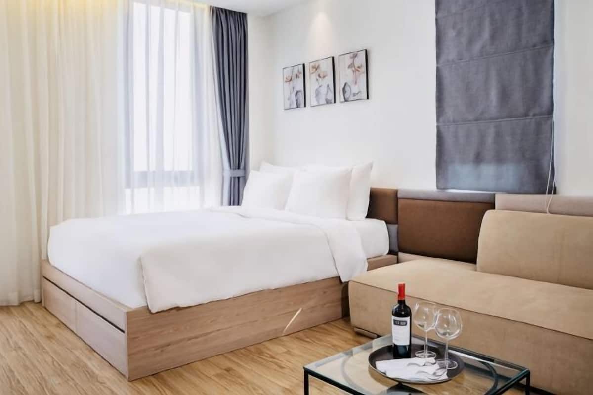Quarto com cama, sofá, mesa de vidro com uma garrafa e copos, e janela com cortinas.