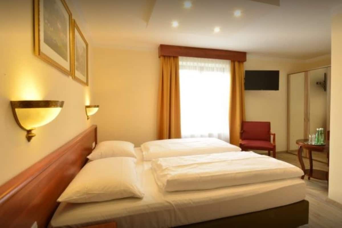 Quarto de hotel com uma cama de casal, duas luminárias fixadas na parede, uma cadeira vermelha, uma mesa pequena e uma janela grande com cortinas.