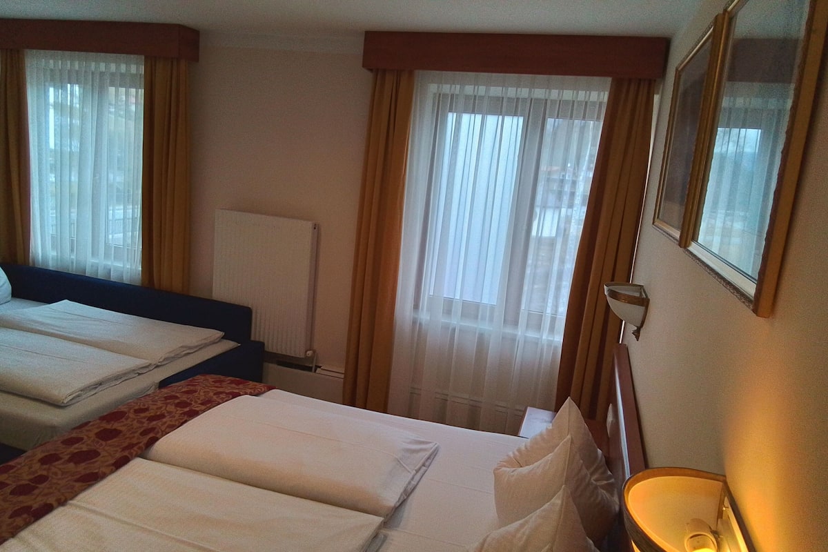 Quarto de hotel com duas camas, um radiador, uma janela com cortinas leves e uma luminária fixada na parede.