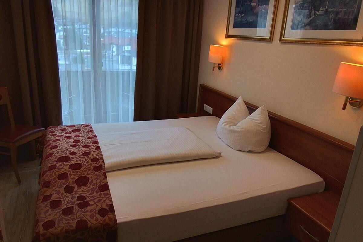 Quarto de hotel com cama, duas luminárias de cabeceira, uma cadeira e janela com cortinas.