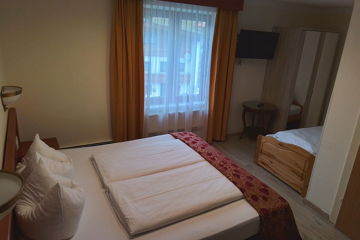 Quarto de hotel com cama, mesa de cabeceira, cadeira, televisão e janela com cortinas.