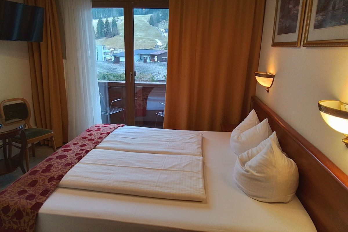Quarto de hotel com cama, escrivaninha, cadeira, televisão e vista para uma varanda com área de estar ao ar livre.