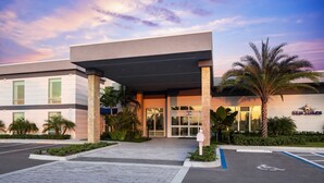 Entrada de hotel com galeria coberta, palmeiras e estacionamento, incluindo vaga para deficientes.