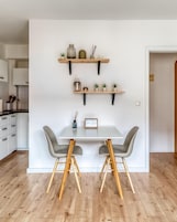 Uma cozinha moderna com armários brancos, bancada de madeira e um espaço de refeições com uma mesa branca e duas cadeiras cinzas.