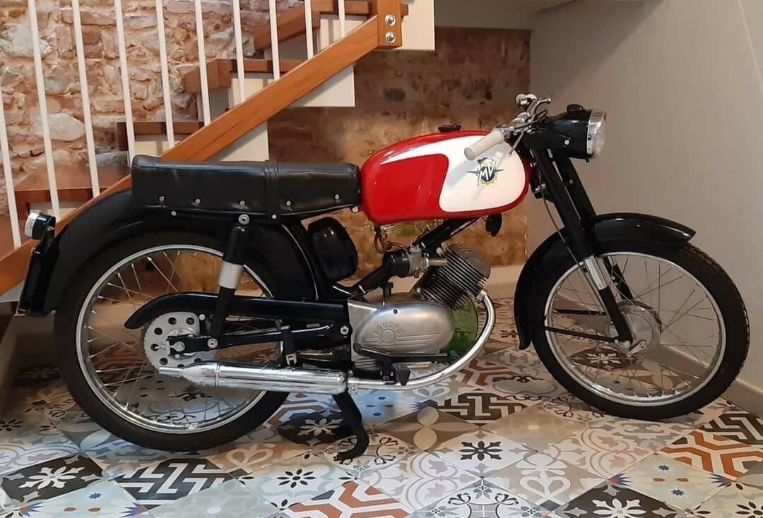 Uma motocicleta vintage com carroceria vermelha e branca, detalhes cromados e assento preto, estacionada em ambiente interno com piso estampado.