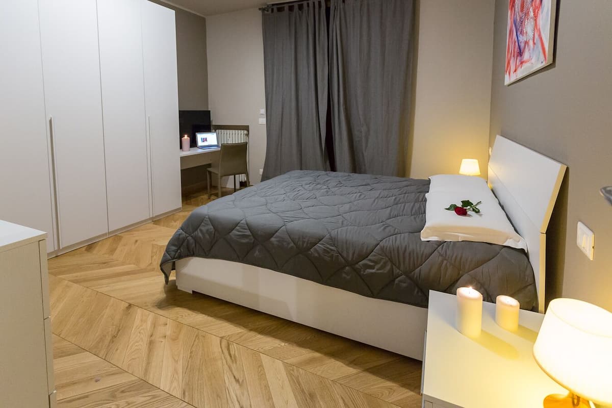 Quarto com cama, mesa de cabeceira com velas, escrivaninha com laptop e cortina cinza.