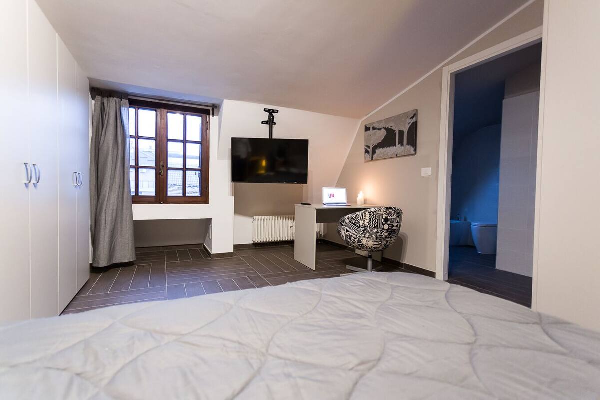 Um quarto com cama, escrivaninha com computador, televisão, janela com cortinas e banheiro.