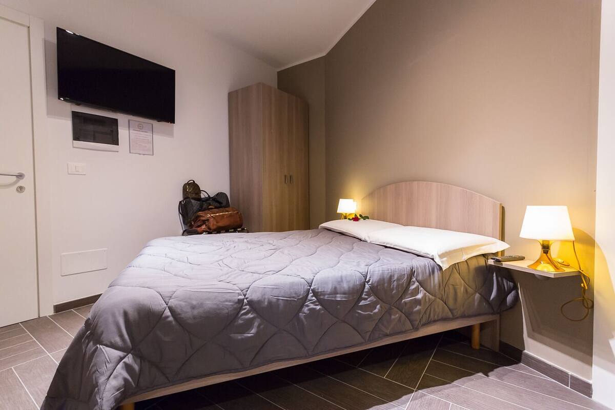 Uma cama bem arrumada com cobertura acolchoada, duas luminárias de cabeceira, uma TV de tela plana na parede e um guarda-roupa de madeira.