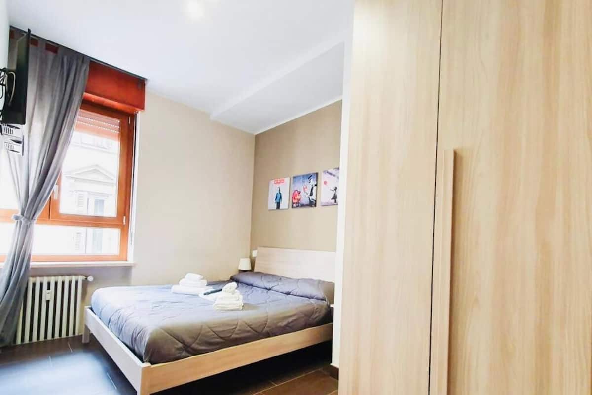 Quarto com cama, radiador, janela com cortinas e guarda-roupa de madeira.