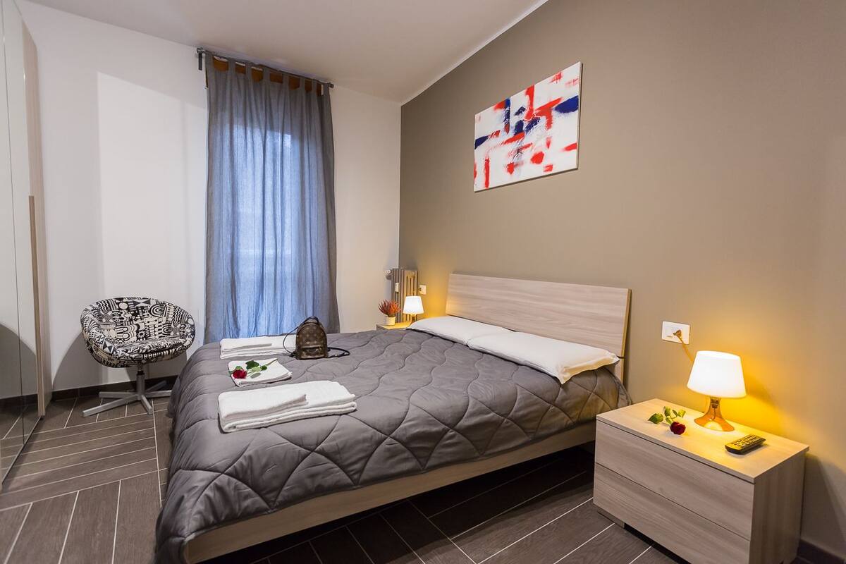 Quarto de hotel moderno com uma cama grande, mesas de cabeceira, uma cadeira e um quadro na parede.