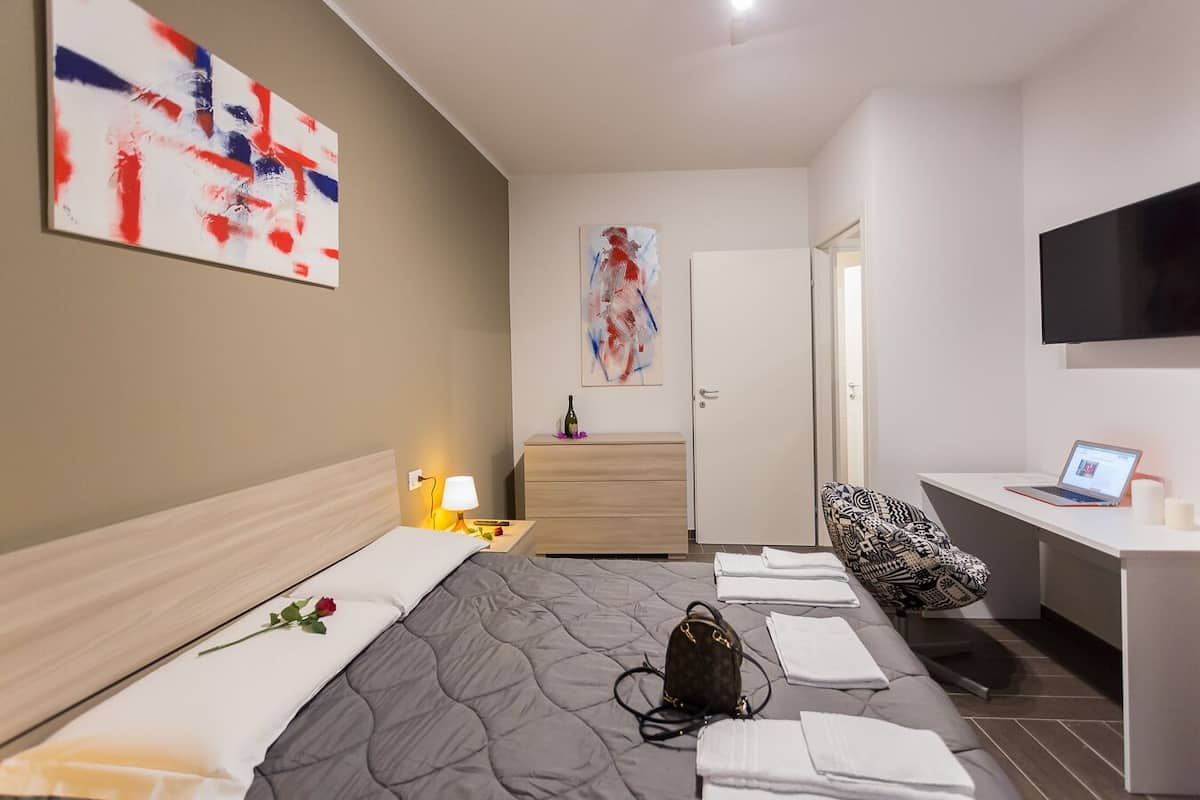 Quarto de hotel moderno com cama, mesa de cabeceira, abajur, obra de arte, escrivaninha, laptop e televisão.