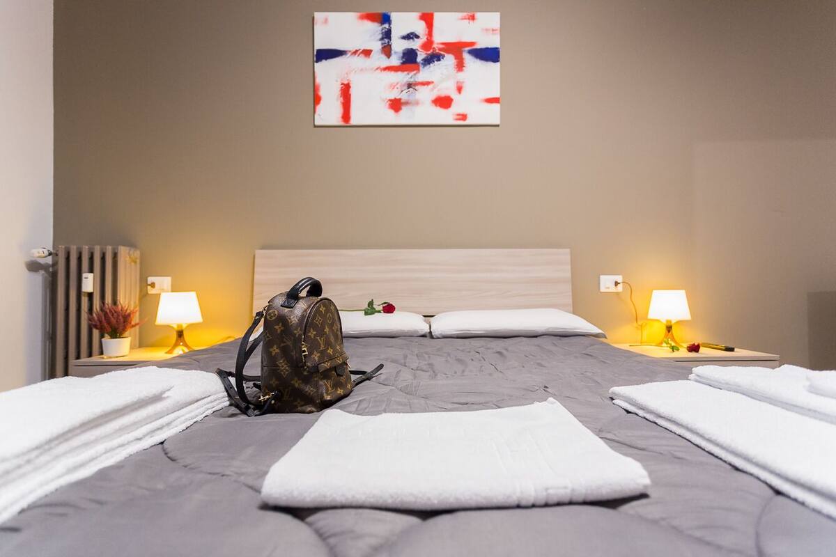 Quarto de hotel com uma cama grande, duas luminárias de cabeceira, um radiador, uma mochila Louis Vuitton e uma rosa vermelha sobre o travesseiro.