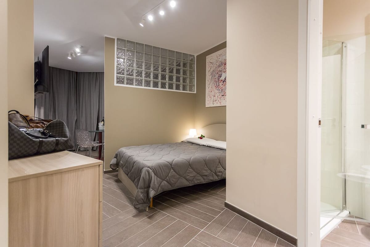 Quarto compacto com cama, escrivaninha, cadeira, janela com persianas e box de vidro.