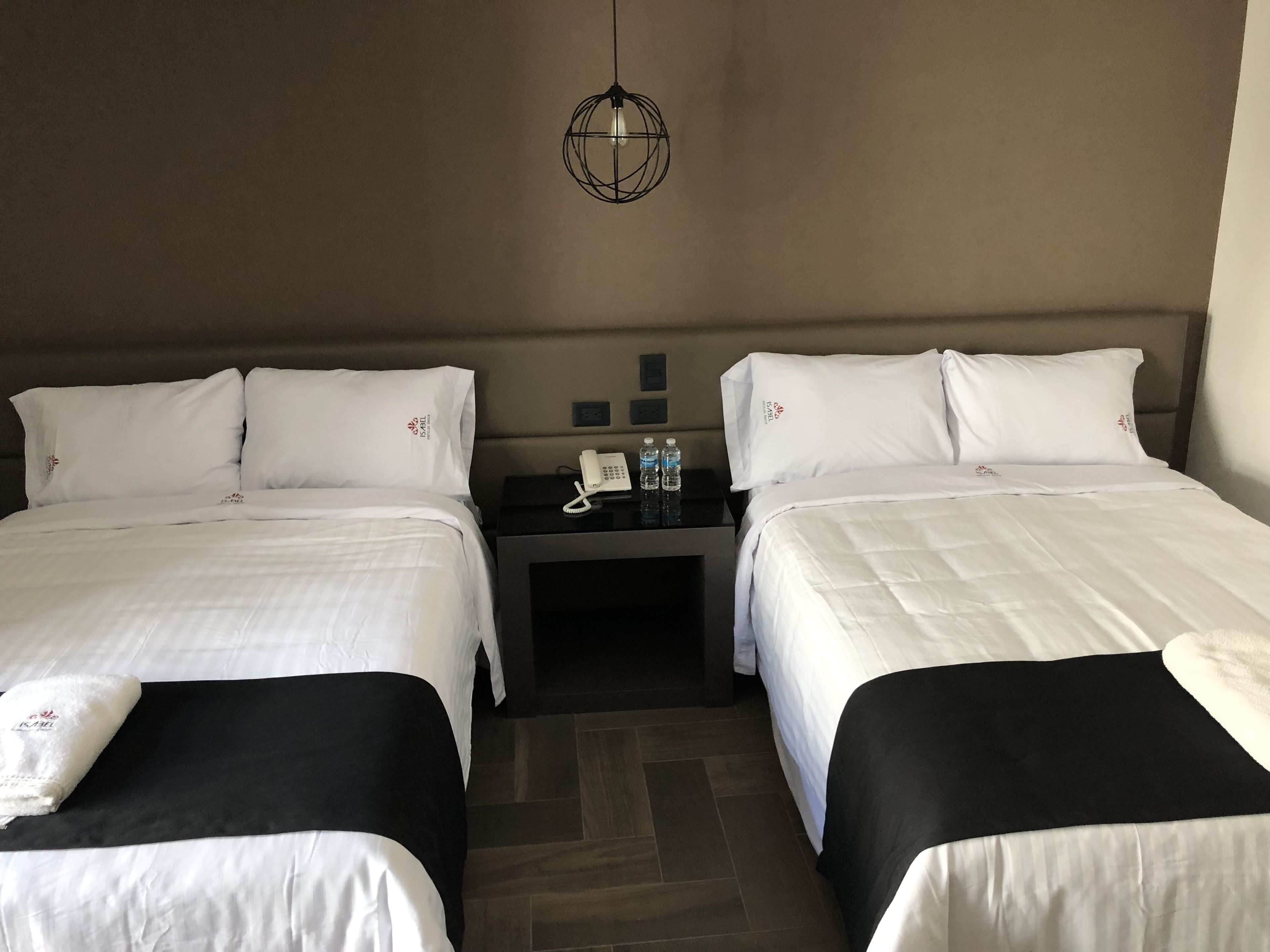 Quarto de hotel com duas camas, cada uma com roupa de cama branca e cabeceiras pretas. Há uma mesinha de cabeceira entre as camas, com um telefone e duas garrafas de água. Um candeeiro globo pendurado no teto.