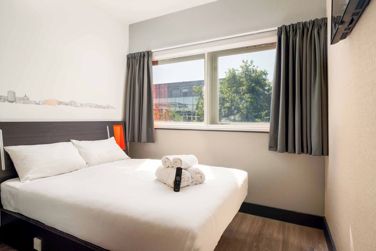 Quarto de hotel com cama, janela com cortinas, controle remoto e toalha sobre a cama.