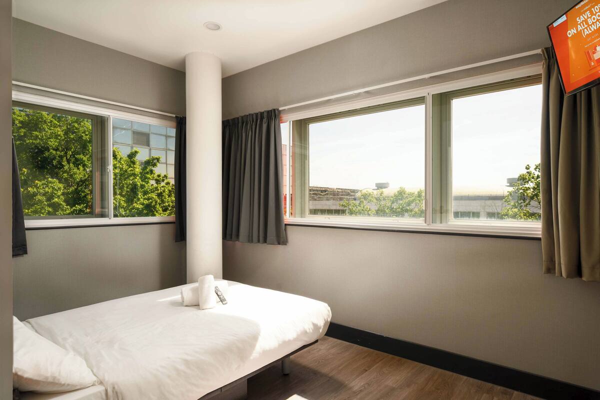 Quarto de hotel com uma cama, janela com cortinas e vista para árvores e edifícios.