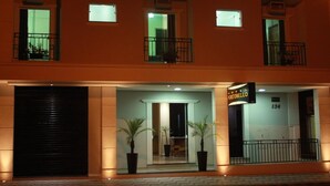 Entrada de hotel com uma persiana fechada, plantas em vasos e uma placa com o nome do hotel.