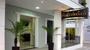 Exterior do Hotel Portobello, com entrada moderna, duas palmeiras em vasos e uma placa.