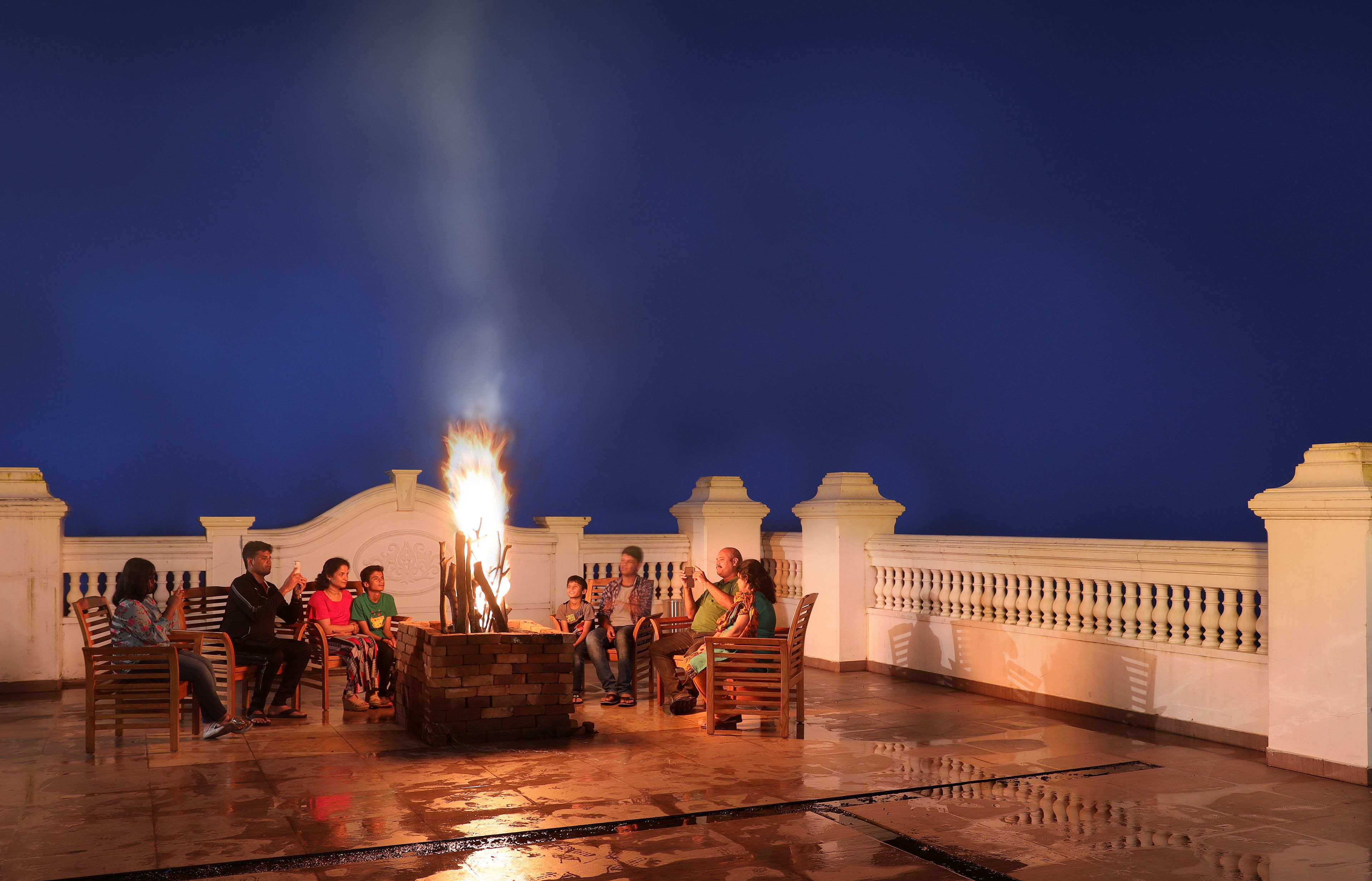 Um grupo de pessoas sentadas ao redor de uma fogueira em um terraço no telhado, com balaústres brancos e céu noturno ao fundo.