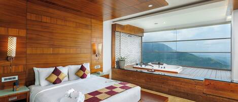 Quarto de hotel moderno com cama grande, painéis de madeira, ampla janela com vista para montanhas, banheiro pequeno e piso de madeira.